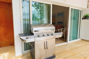 Uma churrasqueira de aço inoxidável com quatro queimadores, forno embutido e queimador lateral, localizada em um ambiente com porta de vidro deslizante que dá para uma área externa.