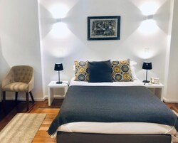 Quarto bem arrumado com cama, duas mesas de cabeceira, uma cadeira e uma obra de arte pendurada na parede.