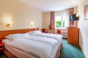 Quarto de hotel com cama de casal, duas luminárias de cabeceira, uma mesinha com uma cadeira, uma TV e uma janela com cortinas.