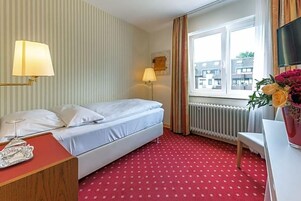 Quarto de hotel com cama, escrivaninha, luminária, radiador e janela com cortinas.