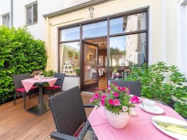 Um pátio externo com cadeiras de vime e uma mesa posta para refeições, uma grande janela com vista para o interior do edifício e plantas em vasos.