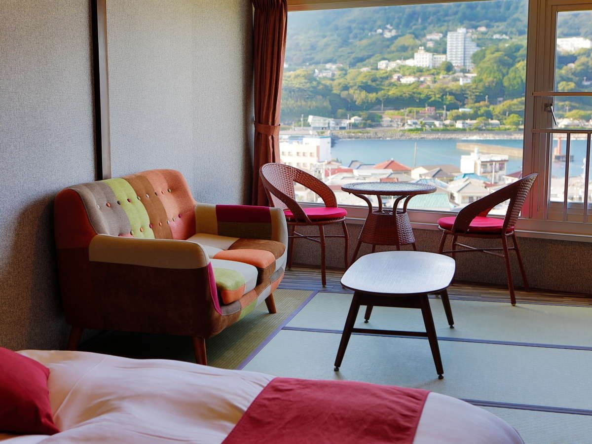 Quarto de hotel com cama, sofá, mesa pequena e duas cadeiras. O quarto oferece vista para um corpo de água e edifícios ao longe.