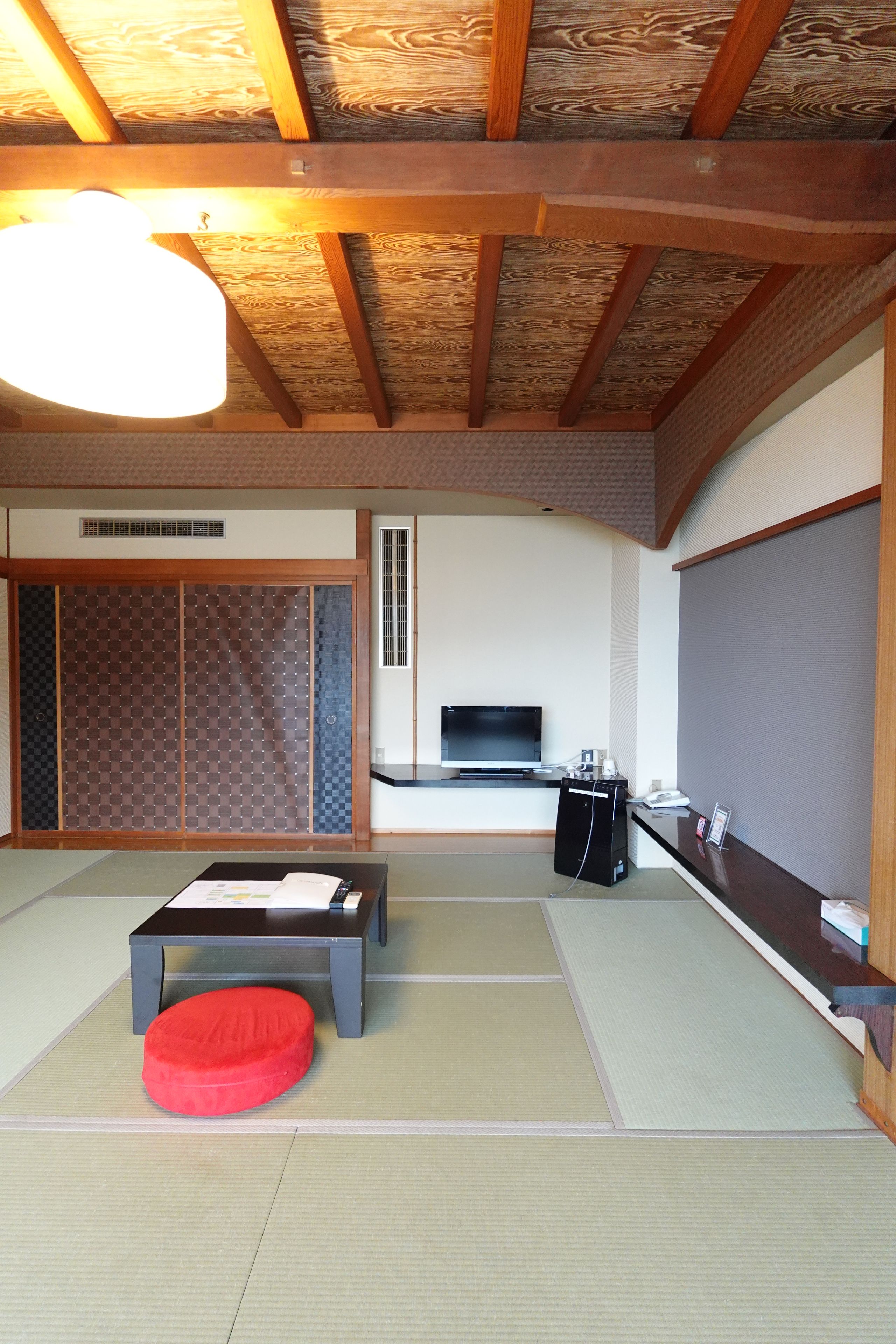 Quarto em estilo japonês tradicional, com tatames, uma mesa baixa, um assento vermelho, uma televisão e um armário preto.