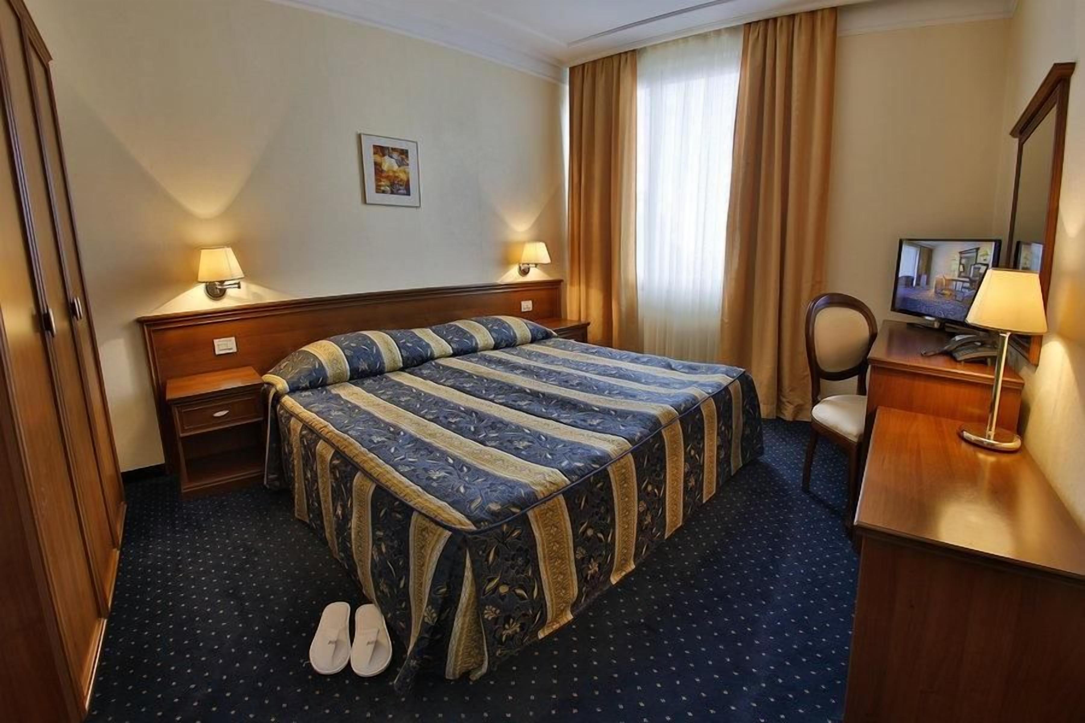 Quarto de hotel com cama, duas luminárias de cabeceira, televisão, escrivaninha com abajur e um par de chinelos no chão.