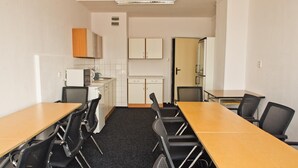 Uma sala de conferências com uma mesa comprida, cadeiras pretas, uma pequena cozinha e armários brancos.