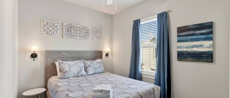 Um quarto com cama, ventoinha no teto, decoração na parede, janela com cortinas e uma pintura na parede.