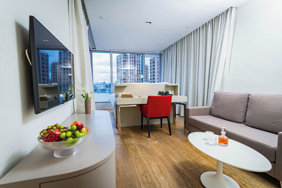 Quarto de hotel moderno com escrivaninha, cadeira vermelha, sofá e uma tigela de frutas no balcão.