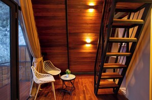 Interior em madeira com escada, janela com cortinas e uma pequena mesa com uma planta.