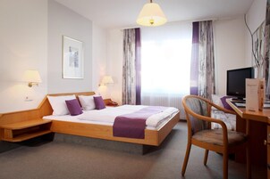 Quarto de hotel com cama, mesas de cabeceira, escrivaninha, cadeira, televisão e cortinas.