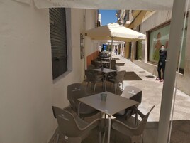 Um café ao ar livre com mesas e cadeiras, um toldo fechado à esquerda e uma pessoa perto da entrada.