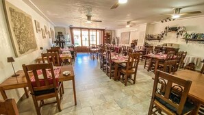Uma sala de jantar ampla, com várias mesas e cadeiras de madeira, piso em ladrilho e ventiladores de teto.