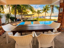 Área de piscina externa com mesa de madeira e cadeiras de vime branco, cercada por palmeiras e vista para o oceano.