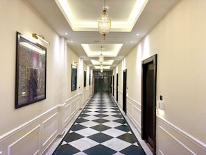 Um corredor de hotel com piso quadriculado, obras de arte emolduradas nas paredes e um candelabro.