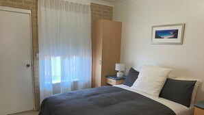 Quarto de hotel com cama, mesa de cabeceira, abajur, quadro pendurado na parede e janela com cortinas leves.