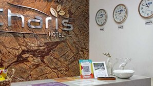 Bancada de recepção com um letreiro 'Welcome to Charis Hotel', relógios mostrando a hora em Jacarta, Sydney e Tóquio, e um código QR para salvar o quarto.