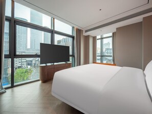 Quarto de hotel moderno com uma cama grande, televisão de tela plana sobre um suporte de madeira e vista para a cidade através de amplas janelas.