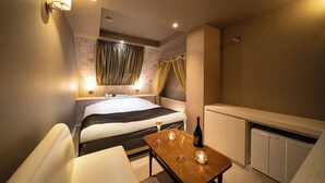 Quarto de hotel com cama, mesa de cabeceira com garrafa e copo, pequena mesa com vela e minibar.