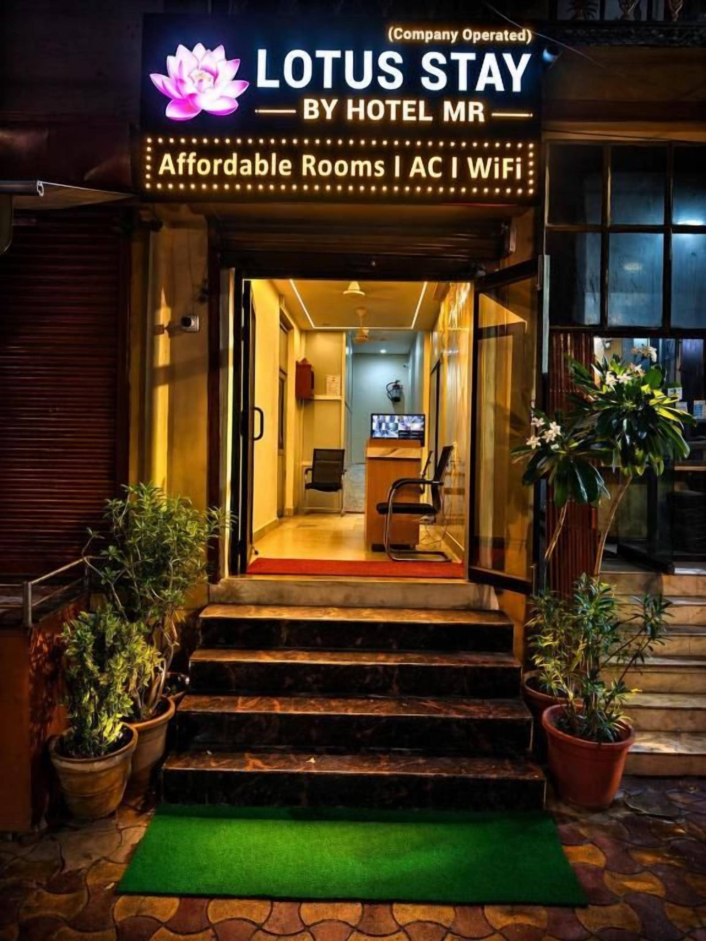 Entrada de um hotel com uma placa indicando 'Lotus Stay by Hotel MR' e mencionando 'Quartos Econômicos | Ar Condicionado | WiFi'.