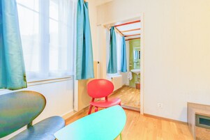 Um quarto com piso de madeira, uma cadeira vermelha, uma cadeira azul e uma janela com cortinas azuis.
