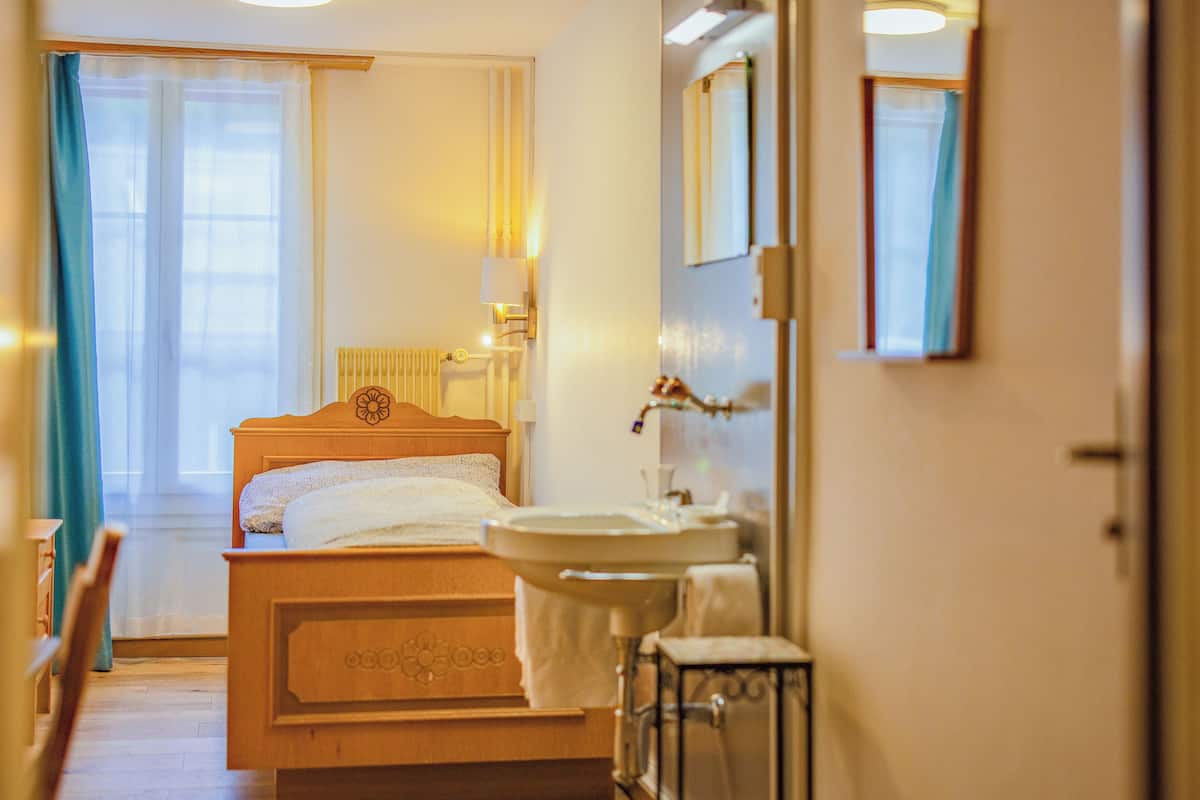 Quarto com cama, mesa de cabeceira, janela com cortinas e banheiro com pia e espelho.