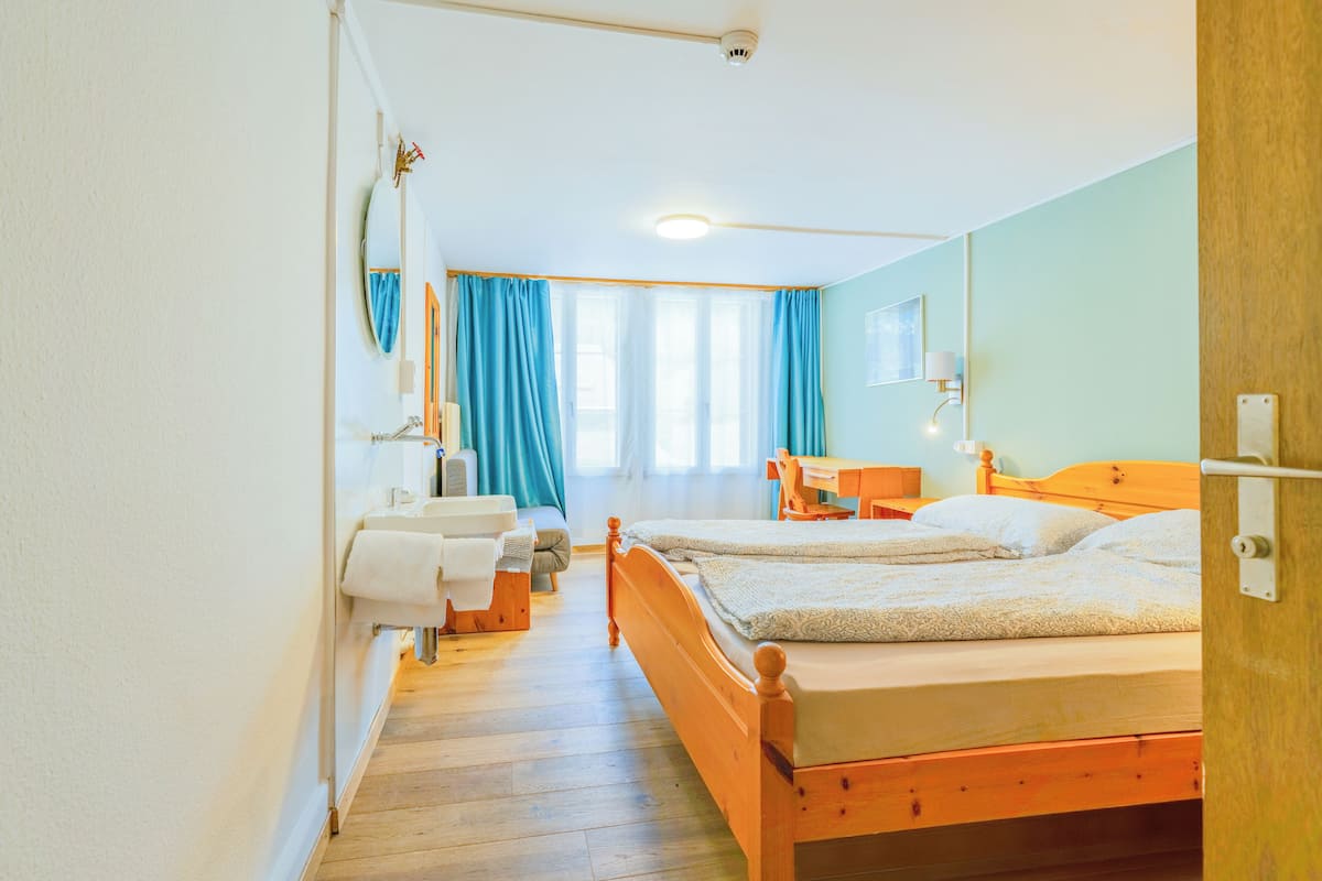 Quarto de hotel com cama de madeira, uma escrivaninha, uma cadeira, um espelho e uma janela com cortinas.