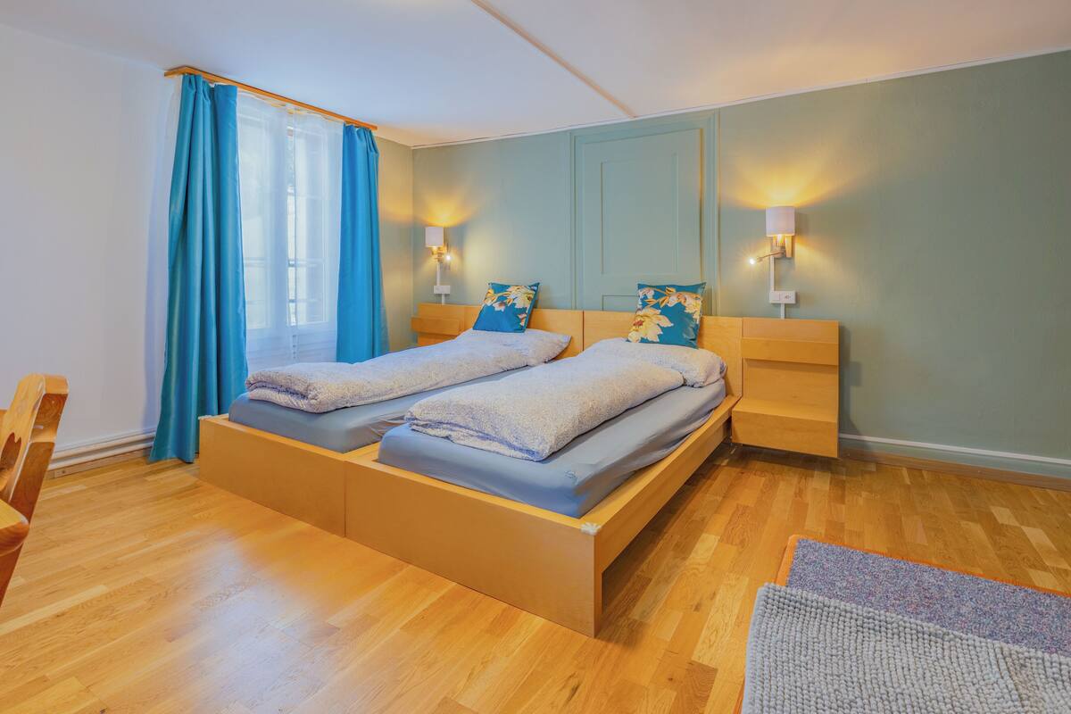 Uma cama de casal com estrutura de madeira, duas mesas de cabeceira com luminárias, uma janela com cortinas azuis e piso de madeira.
