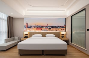 Quarto de hotel com uma cama grande, um sofá, vista para a cidade e uma janela ampla.