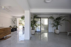 Um lobby moderno com balcão de recepção em madeira, plantas em vasos grandes, ventilador de teto e porta de vidro que dá para o exterior.