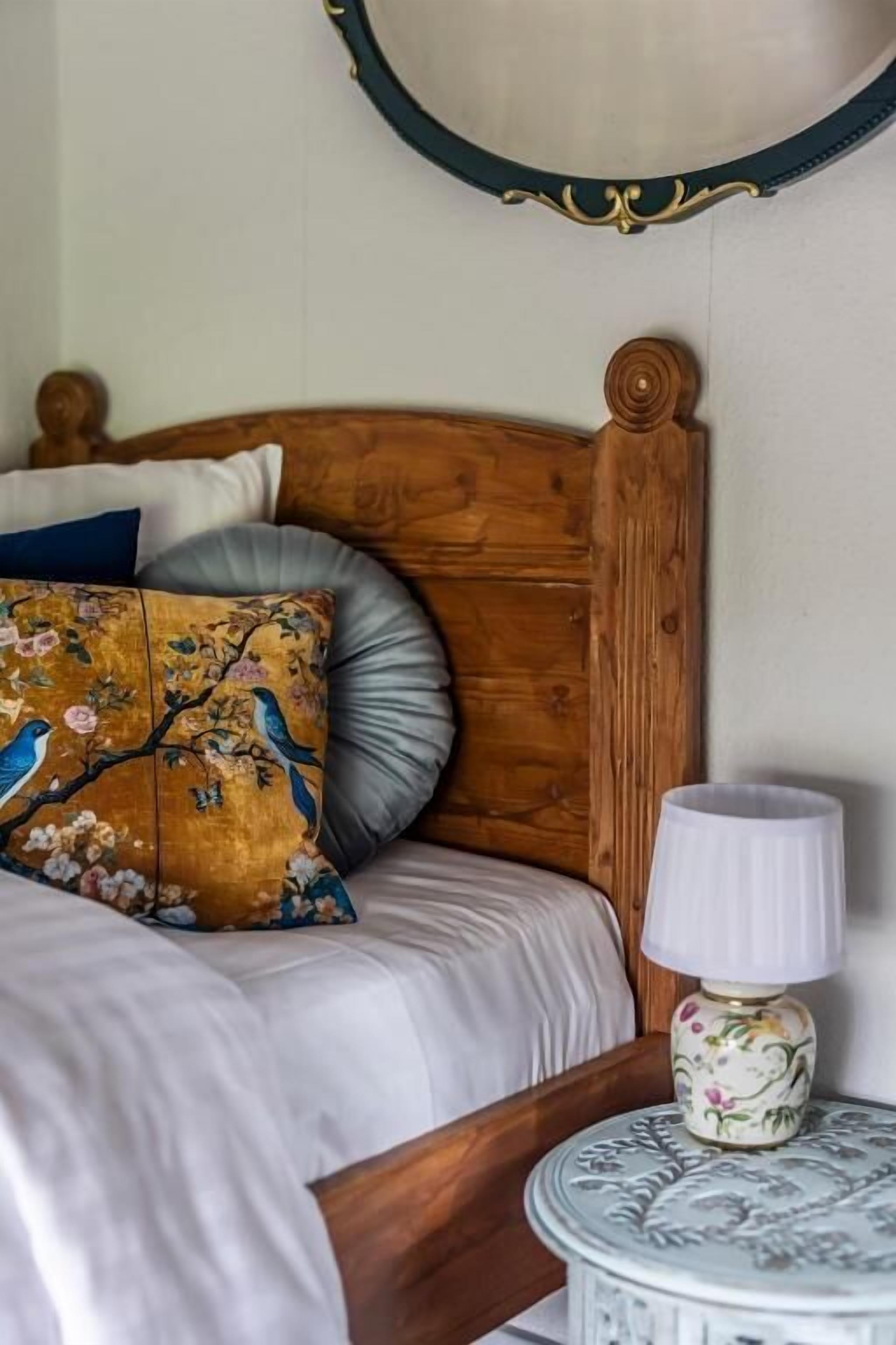 Uma cama de madeira com travesseiro floral, uma mesa redonda com abajur e um espelho na parede.