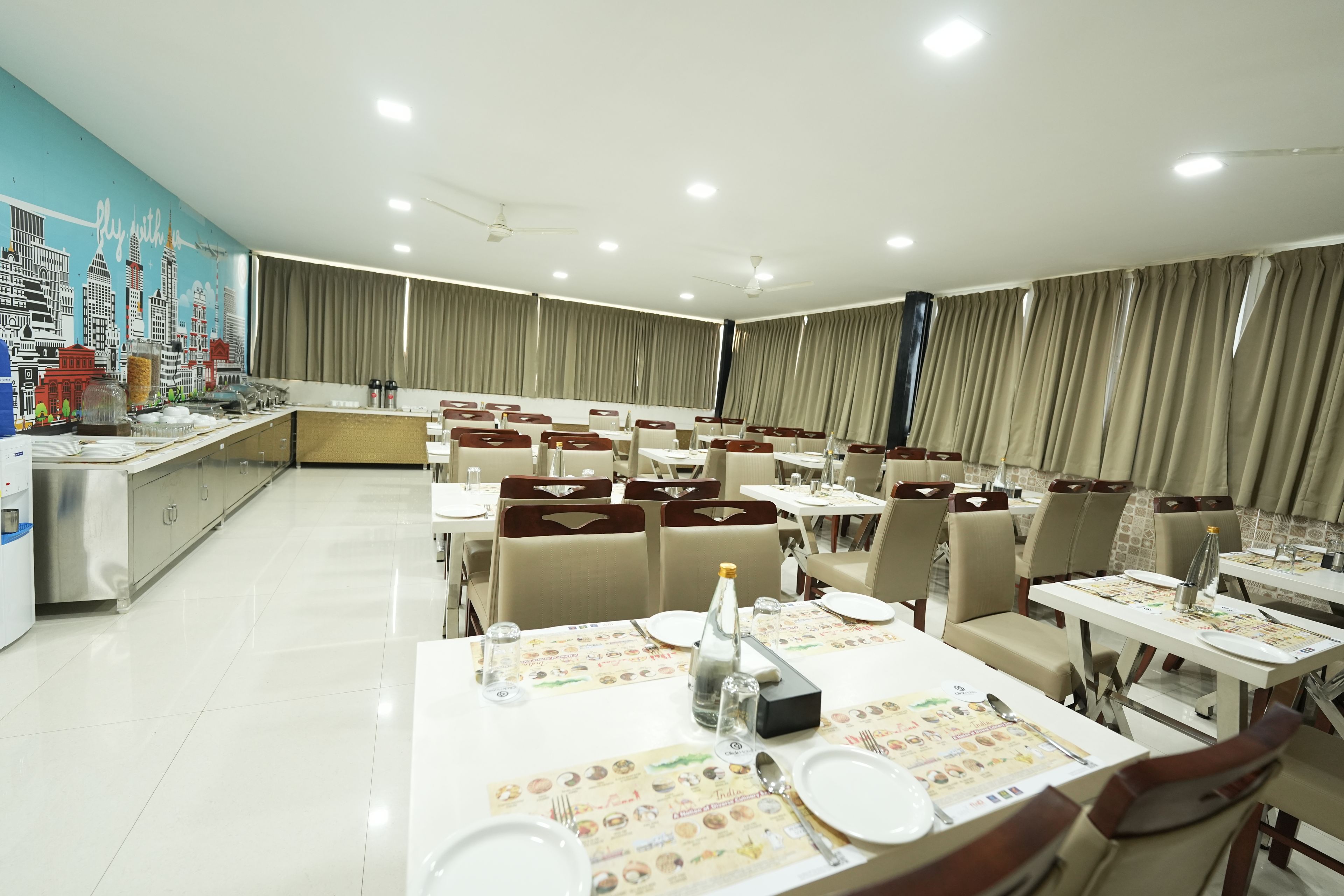 Área de refeições com mesas preparadas para o serviço, cozinha e um mural na parede.