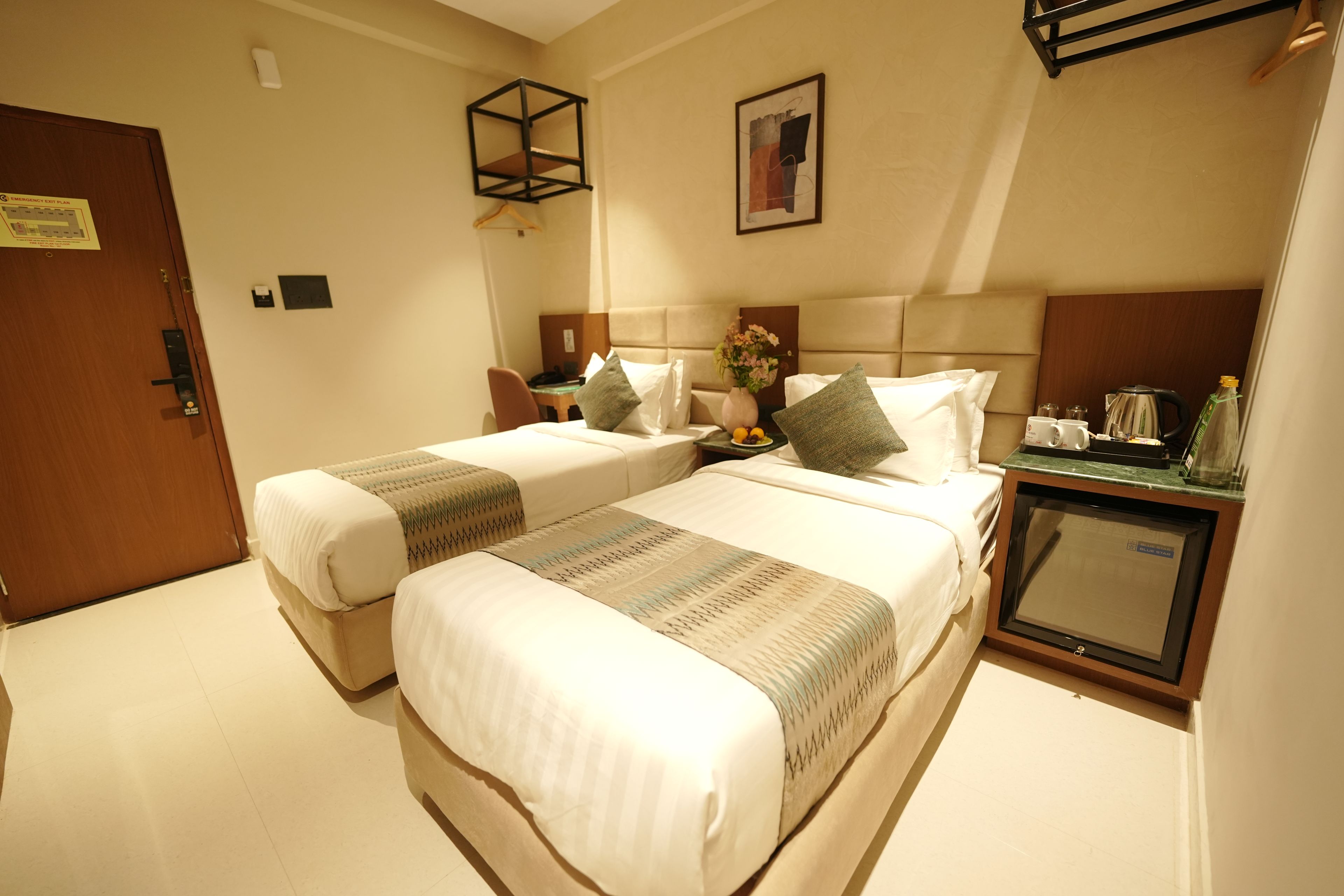 Quarto de hotel com duas camas, uma escrivaninha, um pequeno frigorífico e uma prateleira na parede.