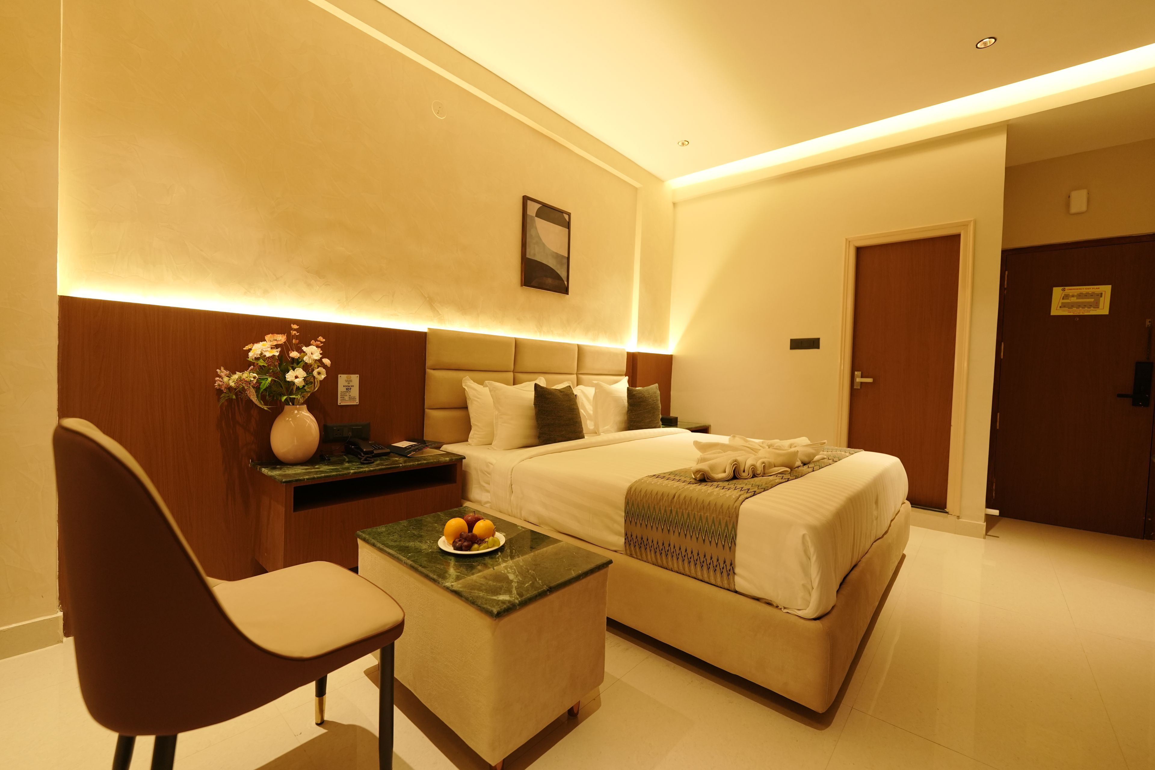Quarto de hotel com cama, mesa de cabeceira, cadeira e uma pequena mesa com frutas.