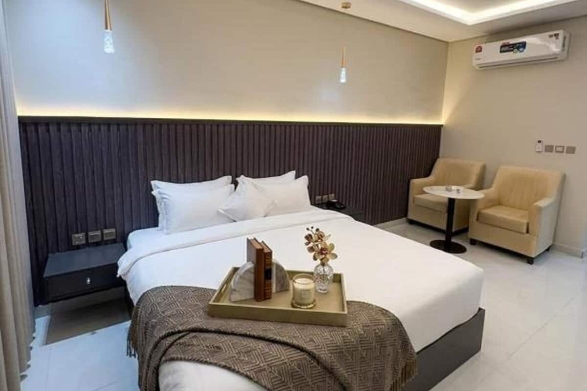 Quarto de hotel moderno com uma cama grande, mesas de cabeceira, um sofá pequeno e um teto com iluminação geométrica.