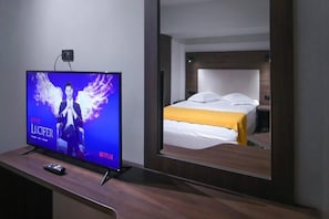 Quarto de hotel com cama, televisão exibindo 'Netflix Lucifer' e controle remoto sobre uma mesa de madeira.