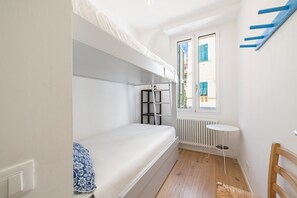 Quarto compacto com uma cama beliche, uma pequena mesa redonda, uma cadeira, radiador e janela com persianas.