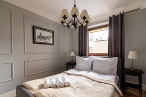 Quarto de hotel com cama, candelabro, quadro emoldurado, janela com cortinas e mesa de cabeceira com abajur.