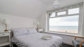 Quarto com cama, duas mesas de cabeceira, abajur, cadeira e uma grande janela com vista para a praia.