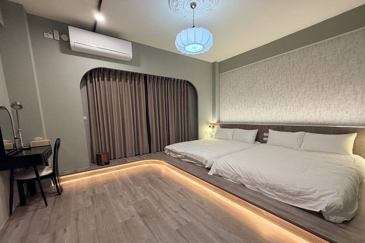 Quarto de hotel moderno com piso de madeira curvado, uma escrivaninha com abajur, cama com roupa de cama branca e luminária no teto.