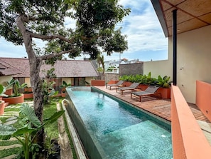 Piscina com espreguiçadeiras, cercada por vasos de plantas e uma árvore.