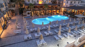Uma área à beira da piscina com espreguiçadeiras e guarda-sóis, piscina e um edifício com vários andares e varandas.