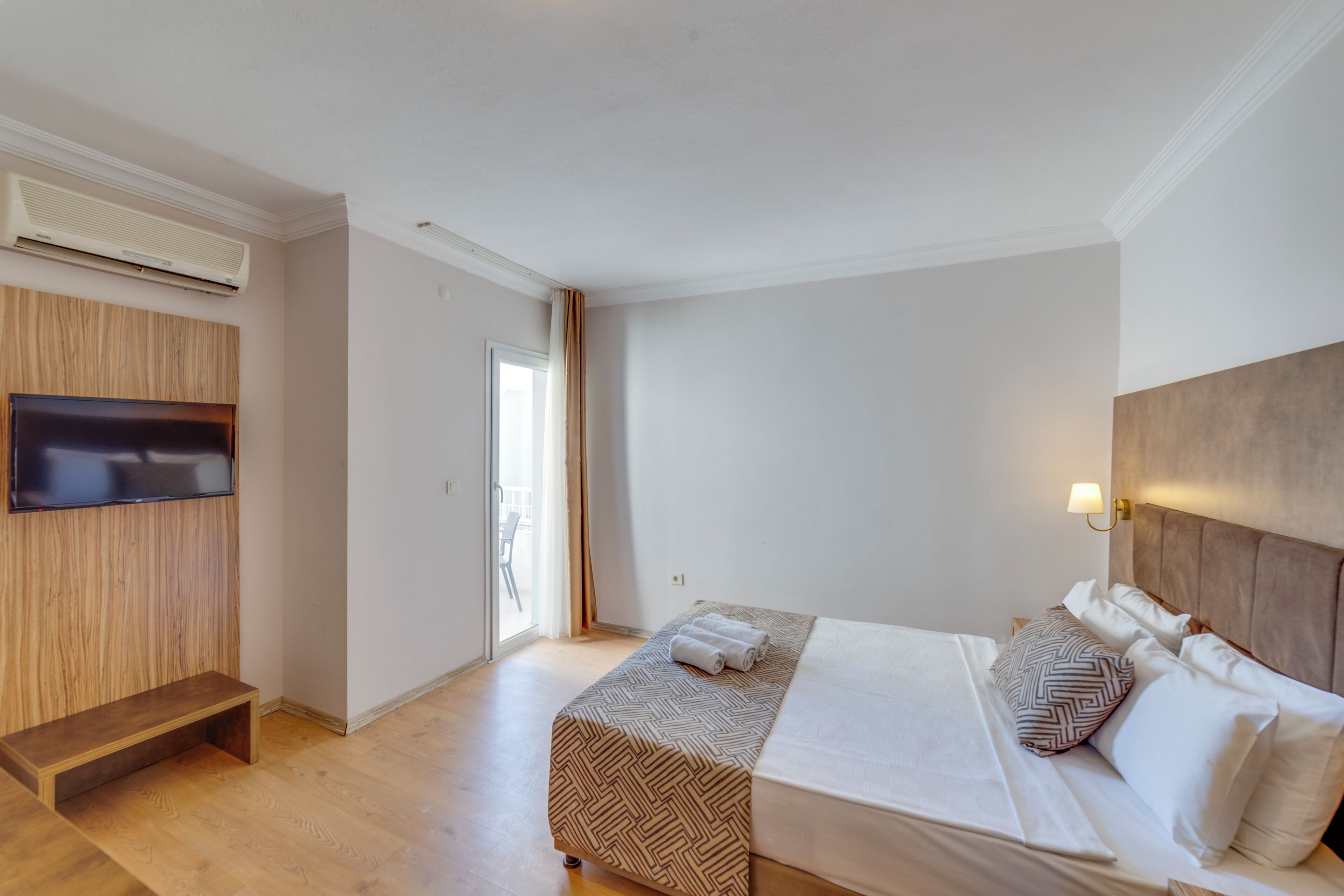 Um quarto com cama, televisão fixada na parede, um banquinho e janela com cortinas.