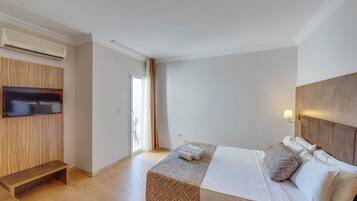 Um quarto com cama, televisão fixada na parede, um banquinho e janela com cortinas.