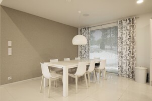 Uma área de refeições com mesa e cadeiras brancas, uma luminária pendente, uma janela com persianas e uma lixeira.