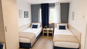 Quarto de hotel com duas camas, uma mesinha, uma lixeira e uma janela com cortinas.