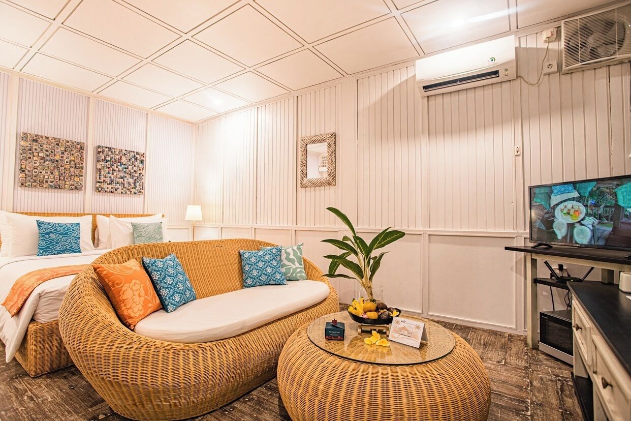 Quarto de hotel com cama, sofá de vime, mesa de centro redonda com uma planta e televisão.