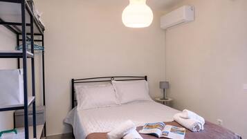 Quarto de hotel com cama, mesa de cabeceira, luminária, prateleira com toalhas e aparelho de ar condicionado.