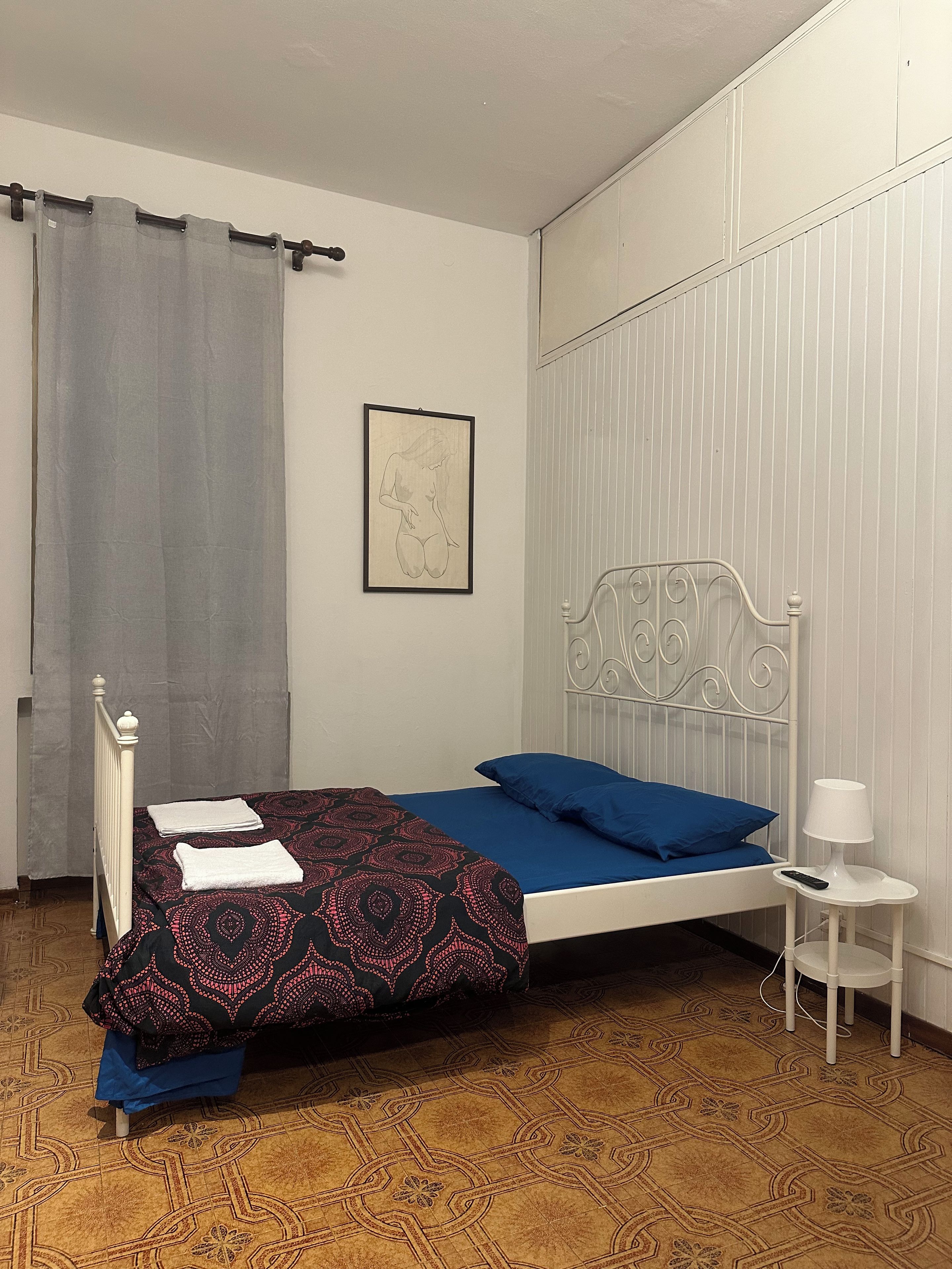 Quarto com cama, mesa de cabeceira, abajur, cortina e quadro na parede.