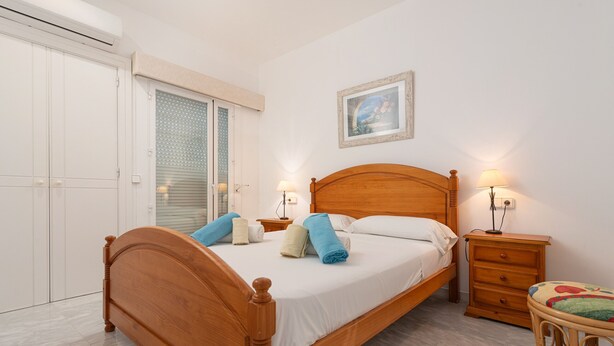 Quarto com cama de madeira, mesa de cabeceira, cômoda, janela com persianas e um quadro na parede.