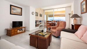Uma sala de estar com televisão, uma mesa de centro, um sofá e um espaço de jantar com cadeiras.