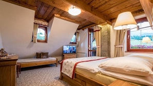 Um quarto aconchegante com teto de madeira, cama com roupa de cama branca, mesa de cabeceira, televisão e janela com cortinas.